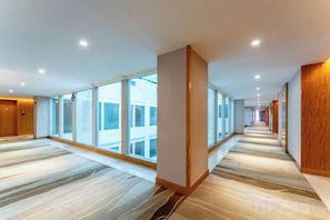 Corredor de hotel com grandes janelas, piso de mármore e painéis de madeira.
