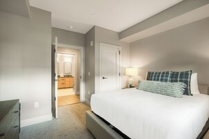 Quarto com cama, mesa de cabeceira, abajur e porta de acesso a um banheiro.