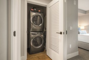 Uma lavanderia com duas máquinas de lavar e secar empilhadas, uma porta branca e um corredor que dá para um quarto.