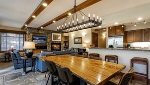 Uma sala ampla com uma grande mesa de jantar de madeira, assentos confortáveis, uma lareira de pedra e uma cozinha com armários de madeira.