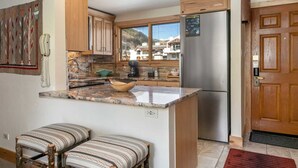 Cozinha com ilha central, armários de madeira, geladeira de aço inoxidável e janela com vista para casas.