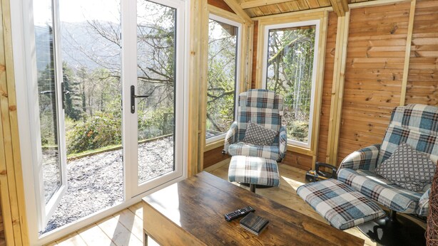 Interior acolhedor de uma cabana, com teto de madeira, uma grande porta de vidro deslizante, um sofá xadrez e uma mesa pequena com um controle remoto.
