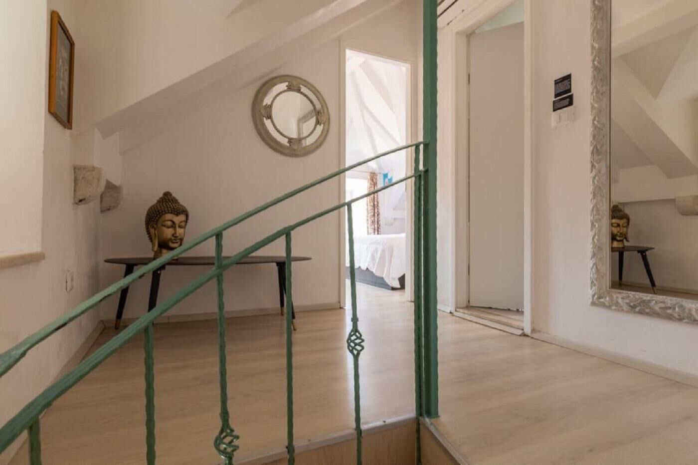 Uma escadaria com corrimãos verdes leva a um quarto com uma estátua de Buda, um espelho redondo e uma cama com roupa de cama branca.