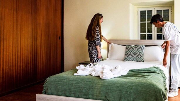 Quarto com guarda-roupa de madeira, cama com colcha verde e janela com persianas.