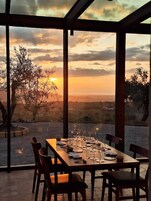 Uma sala de jantar com mesa posta para quatro pessoas, grandes janelas com vista para o pôr do sol sobre a paisagem.