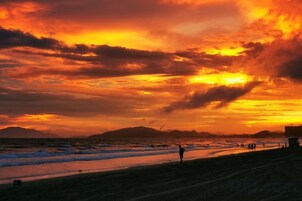 Cena de praia com pôr do sol, silhuetas de pessoas e uma cadeia montanhosa ao longe.