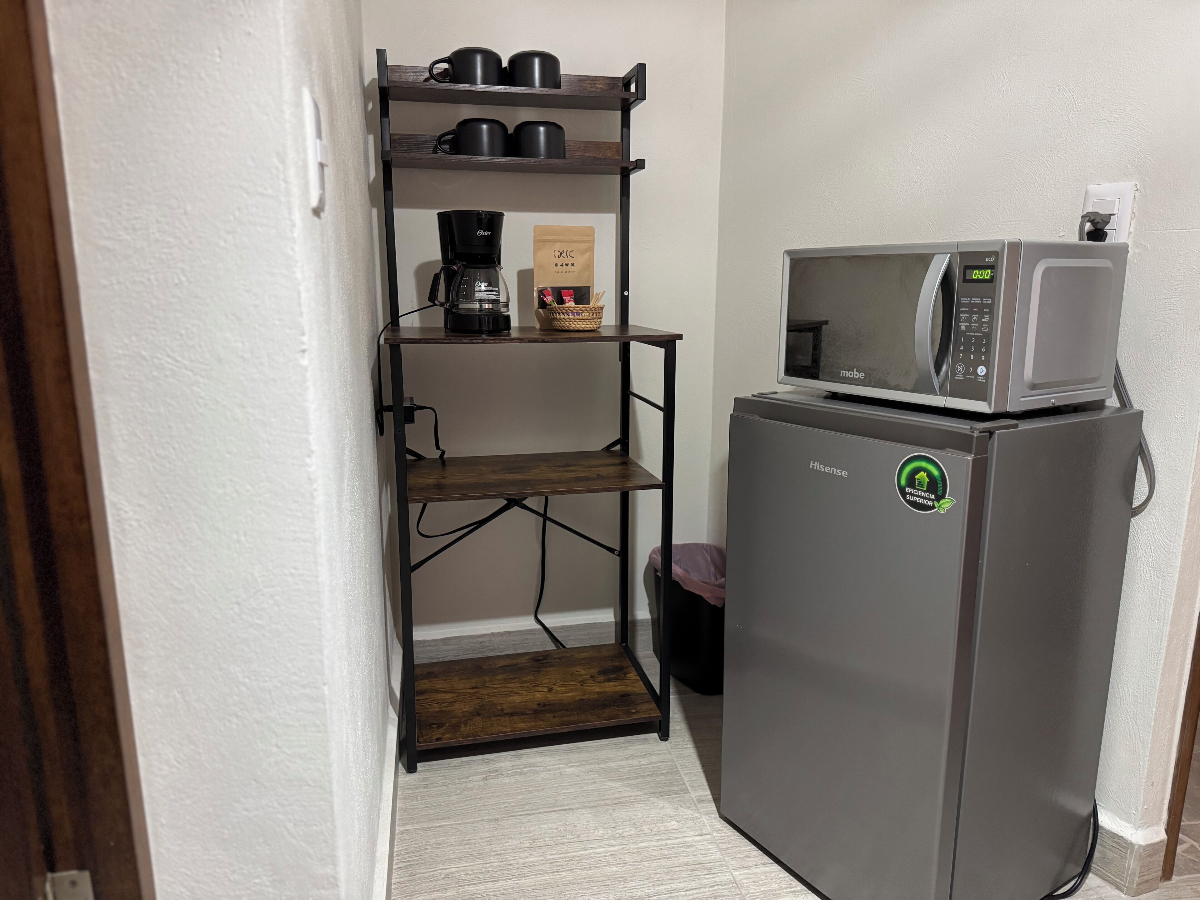 Uma cozinha compacta equipada com uma geladeira Hisense, micro-ondas, cafeteira e uma prateleira com canecas pretas.