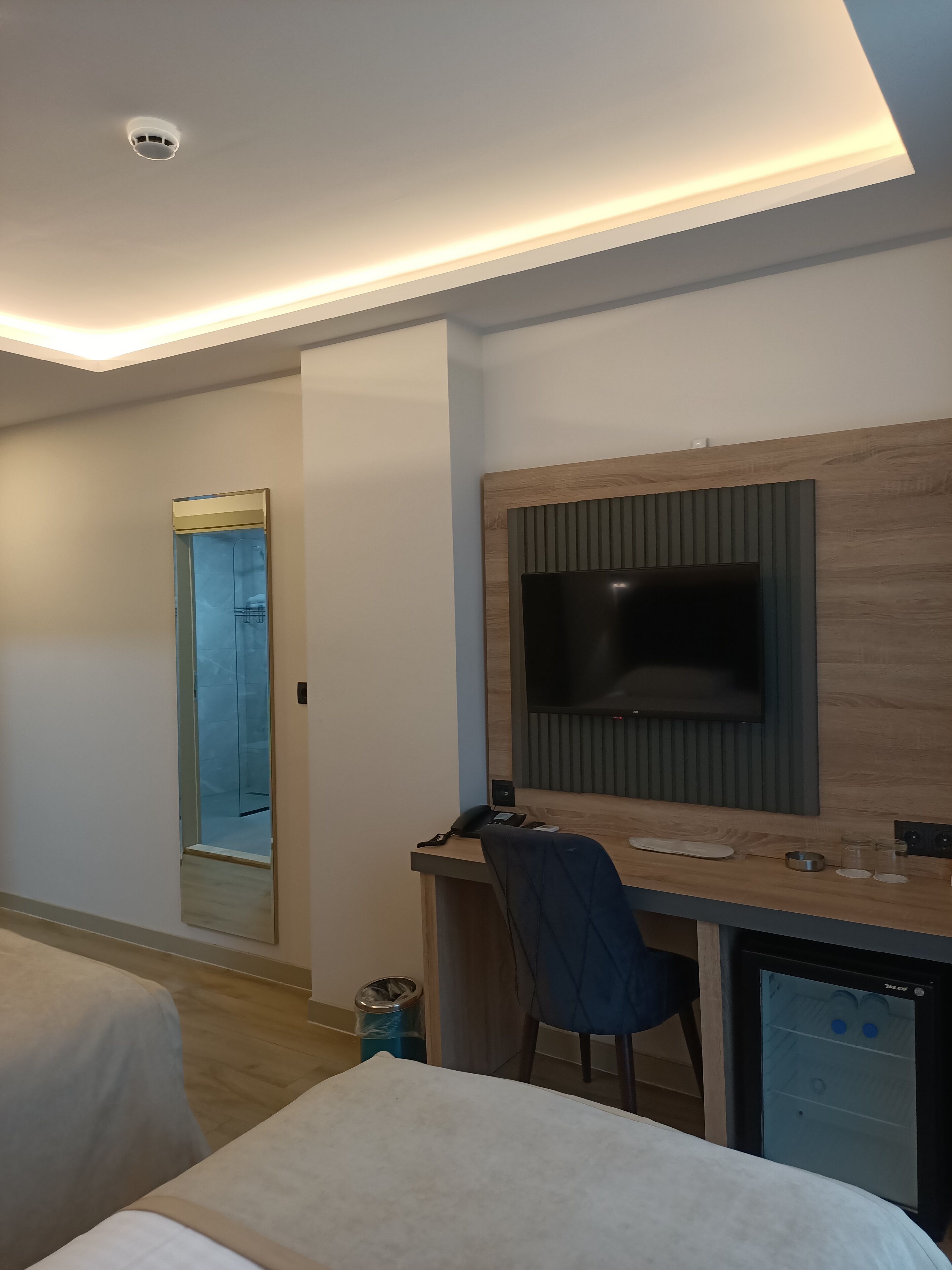 Quarto de hotel com escrivaninha, cadeira, cama, televisão e um pequeno frigorífico.