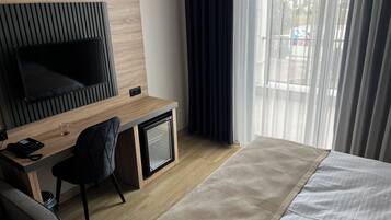 Quarto de hotel com uma escrivaninha de madeira, televisão, frigorífico pequeno, uma cama e vista para o exterior através de cortinas translúcidas.