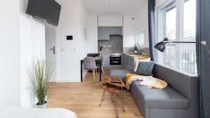 Uma sala de estar moderna com um sofá cinzento, uma mesa de centro e uma televisão na parede. Há uma área de jantar com mesa e cadeiras, e uma cozinha ao fundo.