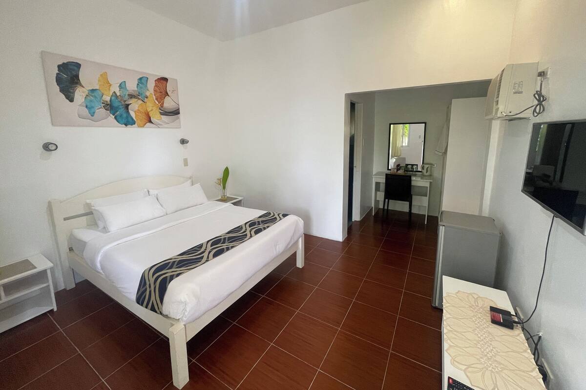 Quarto com cama, escrivaninha, cadeira, televisão e banheiro visível por uma porta aberta.