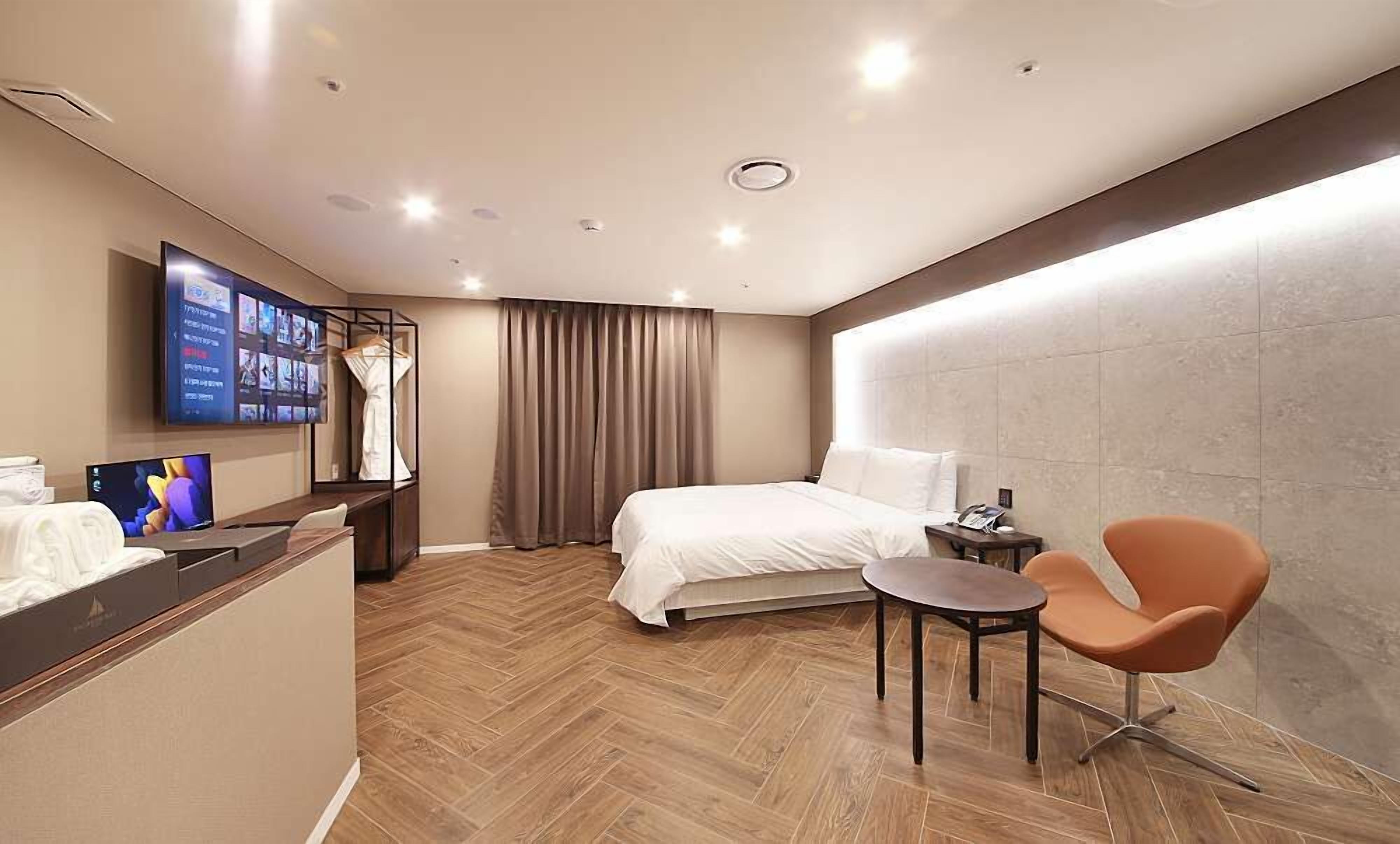Quarto de hotel moderno com cama, escrivaninha com computador, cadeira e televisão fixada na parede.