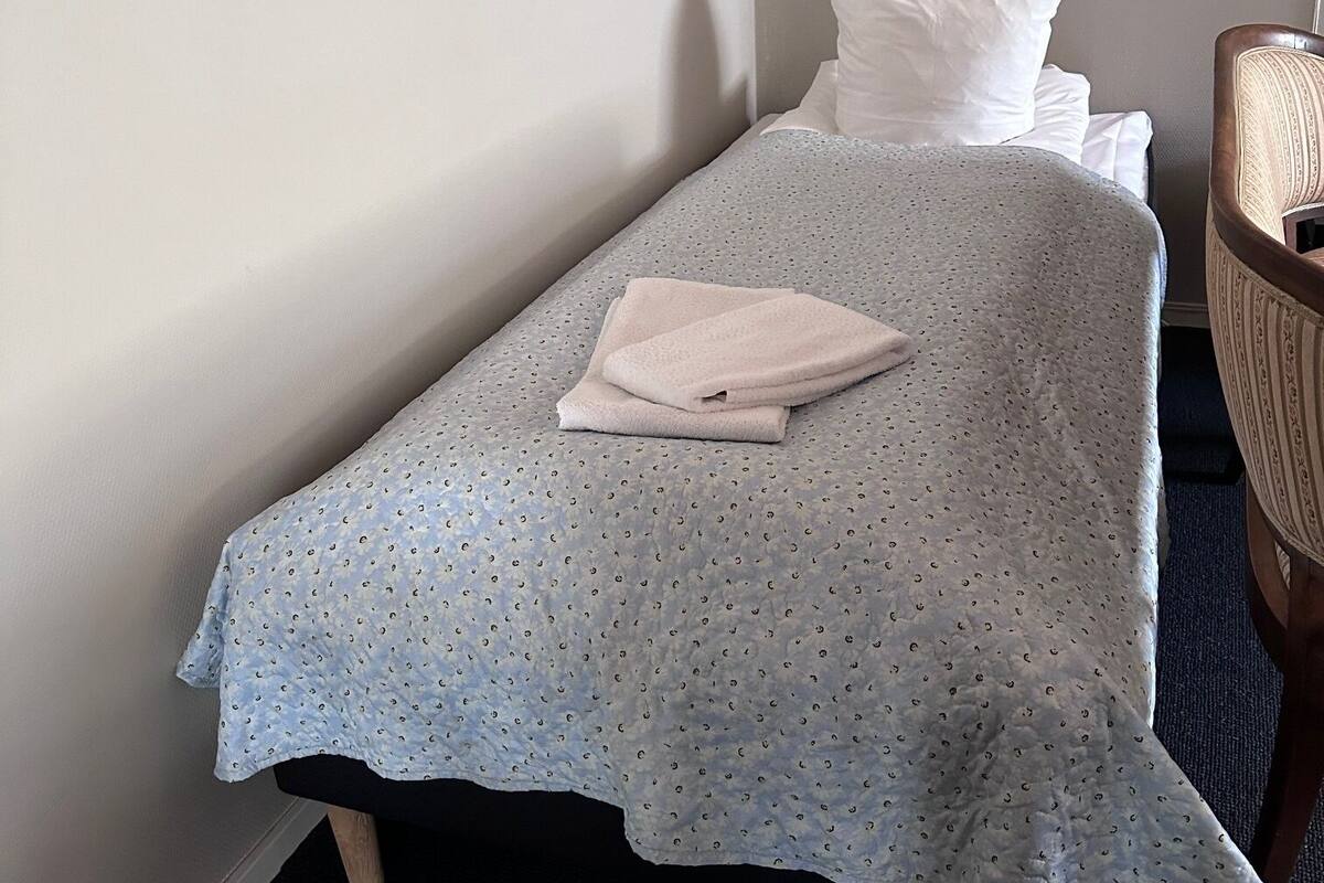 Uma cama pequena e bem arrumada com colcha azul estampada e travesseiros brancos, uma toalha dobrada e uma cadeira de madeira com almofada listrada.