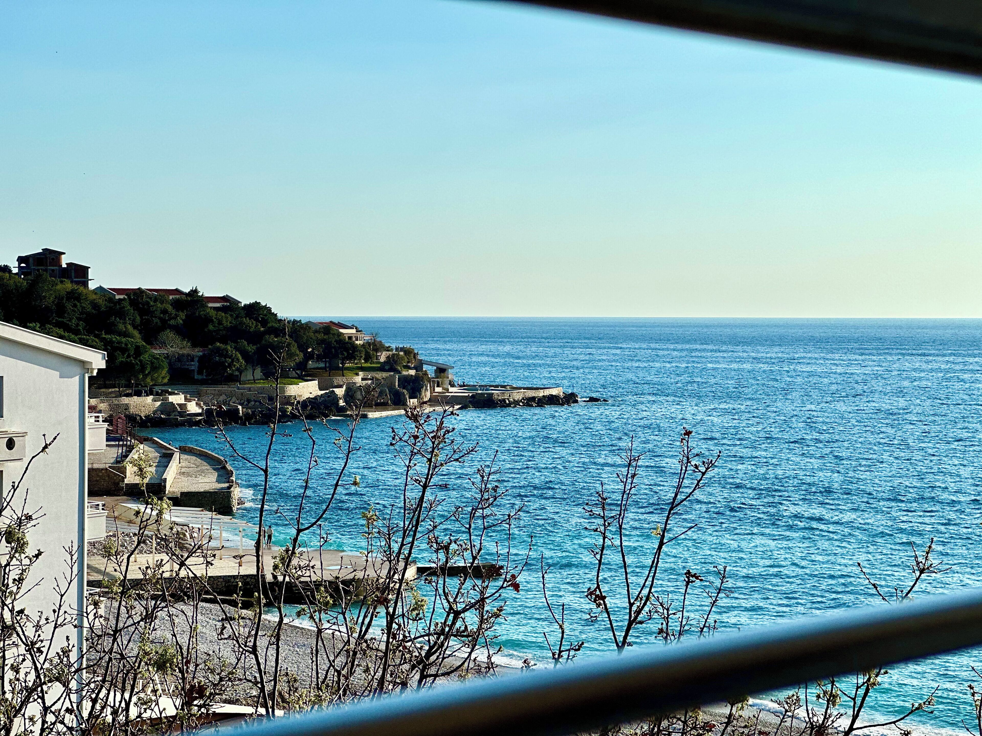 Vista costeira com um cais de pedra, edifícios e mar azul límpido.