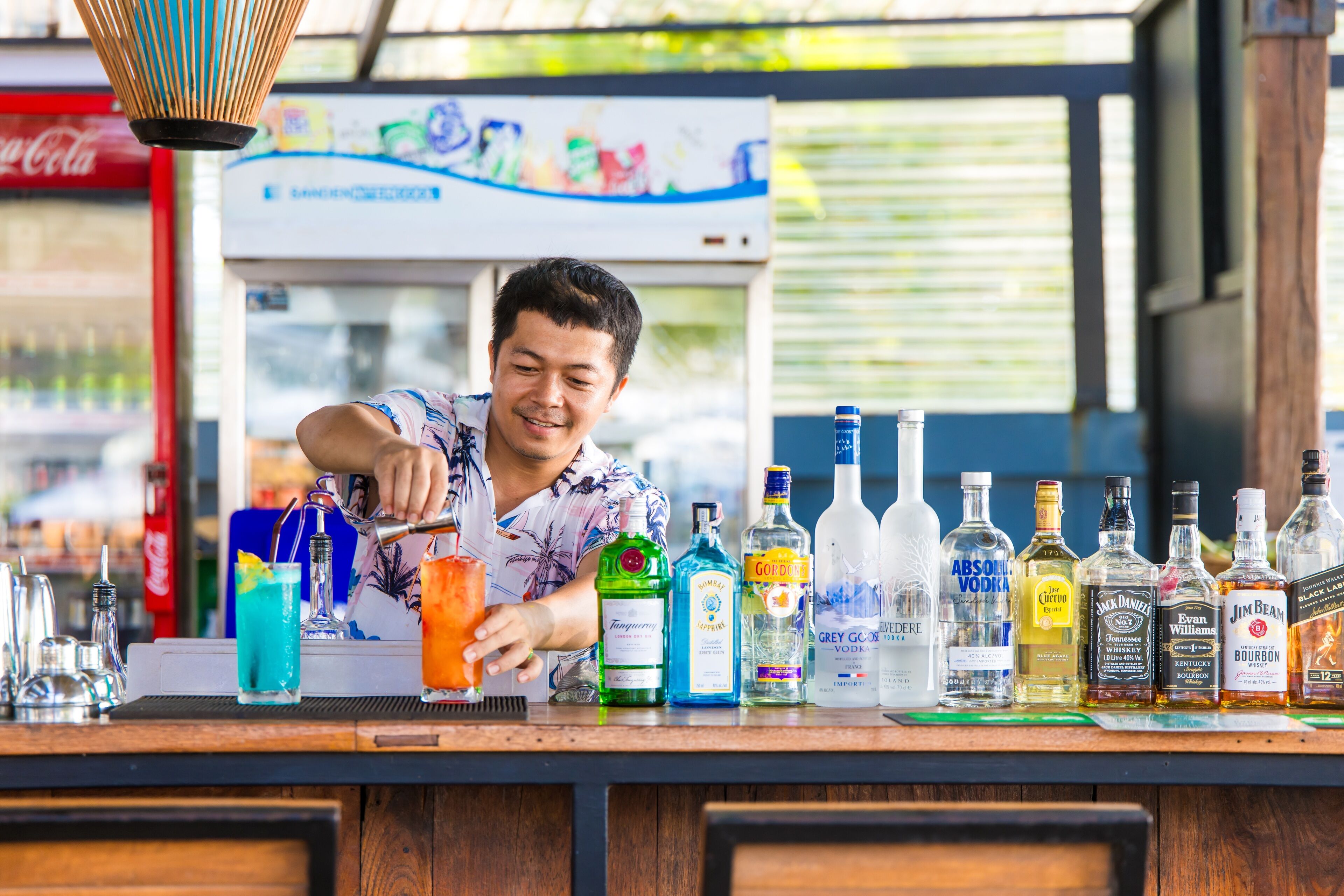 Um bartender prepara bebidas num bar, com diversas garrafas de álcool e um refrigerador de Coca-Cola ao fundo.