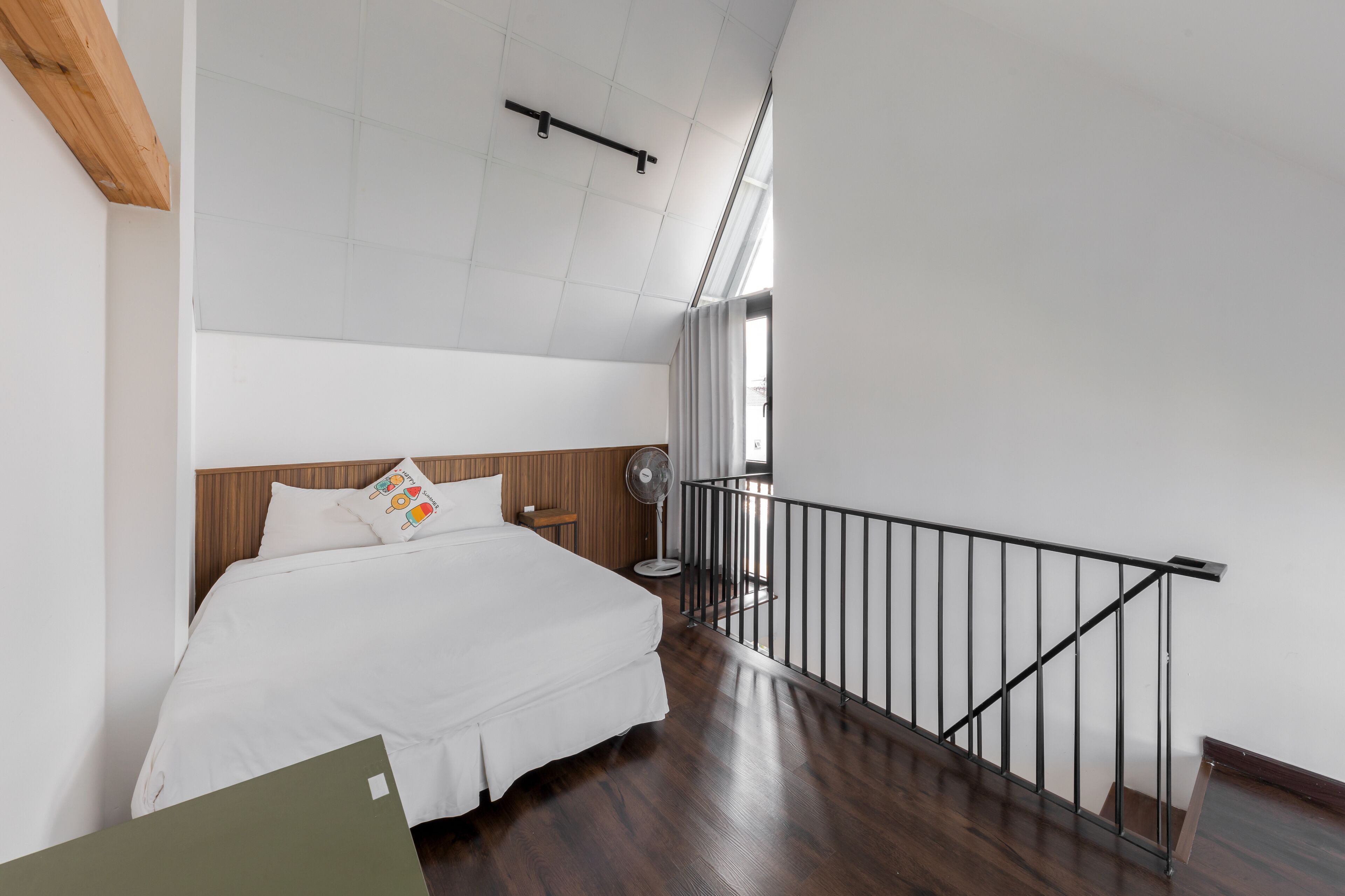 Quarto com cabeceira de madeira, cama com roupa de cama branca, um ventilador e escada com corrimão preto.