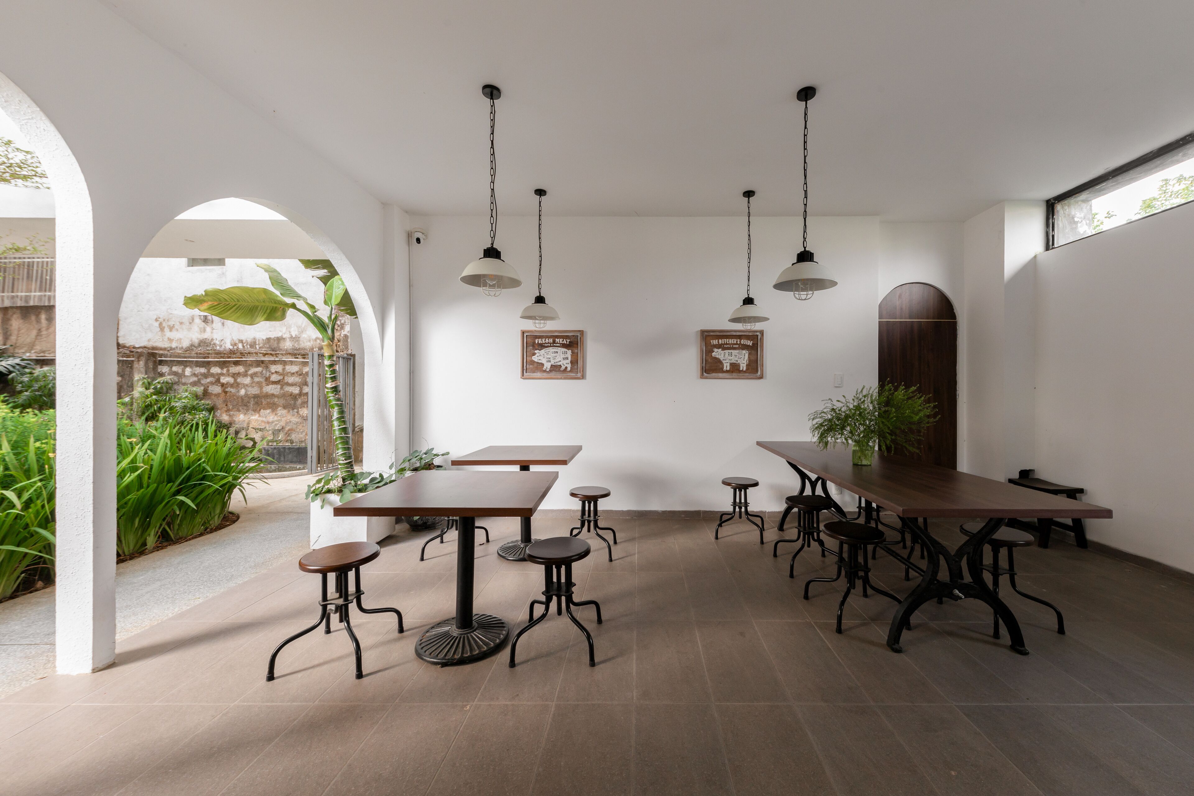 Uma área de refeições com várias mesas e cadeiras, paredes brancas e luminárias pendentes.