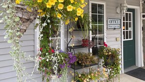 Uma varanda com flores amarelas penduradas, uma placa com a inscrição 'OFFICE Welcome to Green Gables' e diversos vasos de plantas.