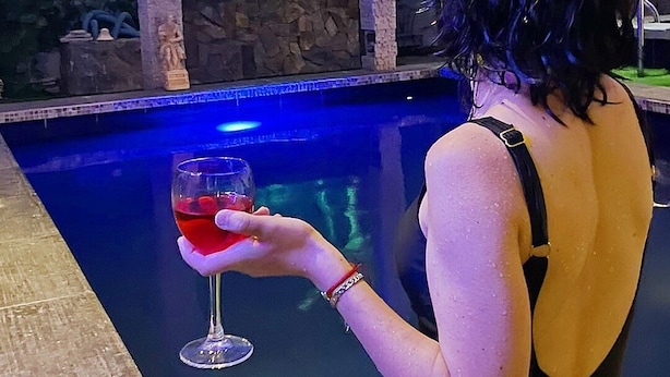Uma pessoa sentada perto de uma piscina, com uma bebida, uma estátua e um muro de pedra ao fundo.