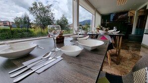 Mesa de jantar posta com tigelas brancas, talheres e copos de vinho, com vista panorâmica para montanhas e vegetação.