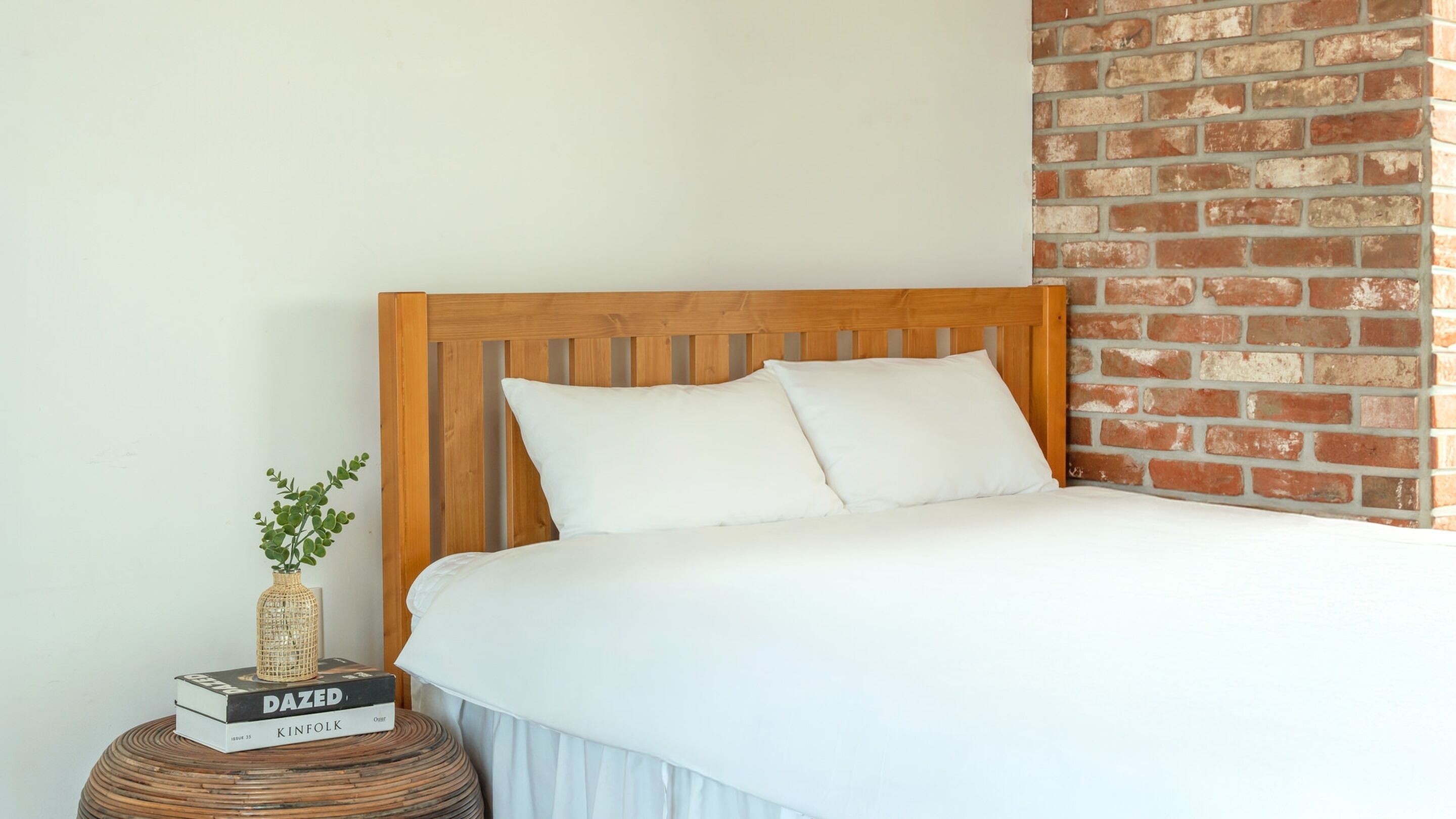 Uma cama com cabeceira de madeira, roupa de cama branca e uma pequena mesa redonda com um livro e uma planta.