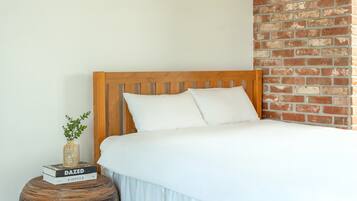 Uma cama com cabeceira de madeira, roupa de cama branca e uma pequena mesa redonda com um livro e uma planta.