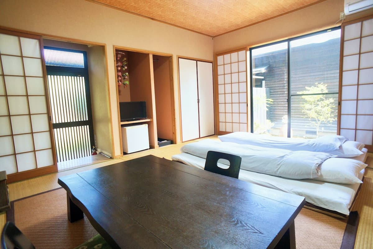 Um quarto japonês tradicional com tatames, uma mesa baixa, uma cama com roupa de cama branca, portas deslizantes e uma televisão.
