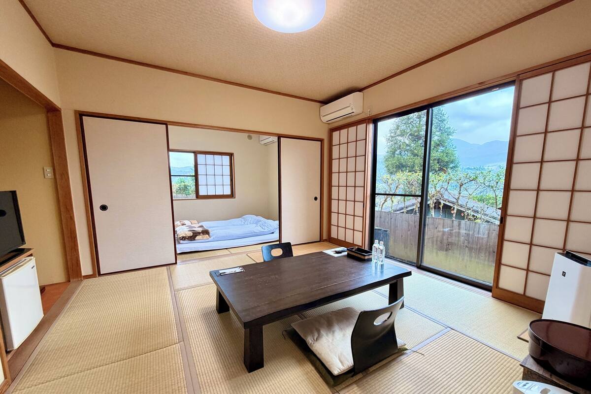 Um quarto japonês tradicional com piso de tatame, uma mesa baixa com almofadas, uma porta deslizante e vista para um jardim através de grandes janelas.