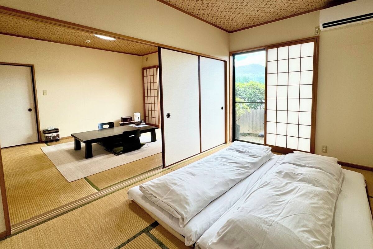 Um quarto japonês tradicional com tatames, futon, mesa baixa com uma cadeira, portas deslizantes e uma grande janela com vista.