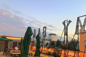 Uma área externa com mesas e cadeiras, guarda-sóis e vista para edifícios ao fundo.