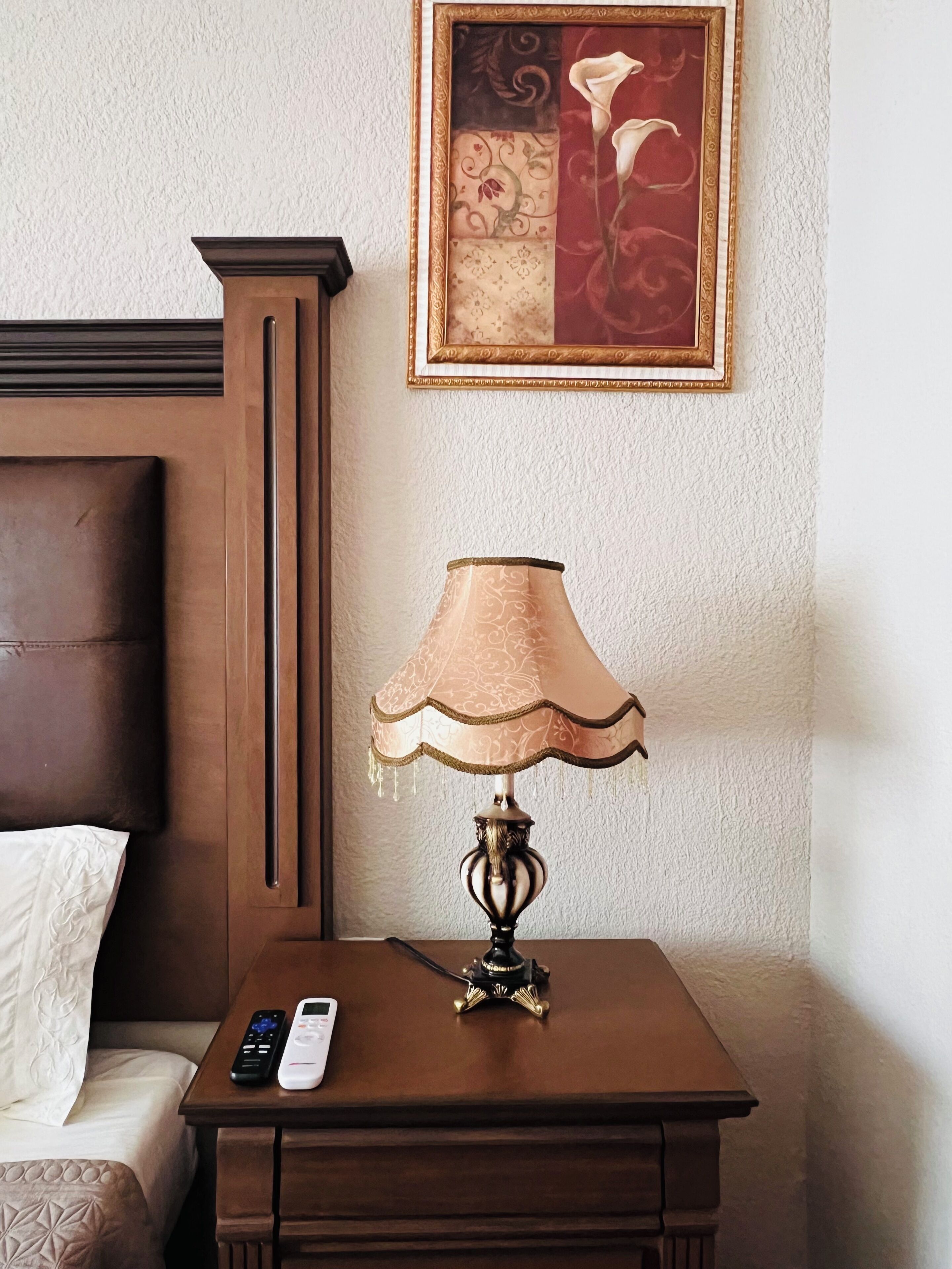 Uma cama bem arrumada com cabeceira, mesa de cabeceira com abajur, controle remoto e um quadro na parede.