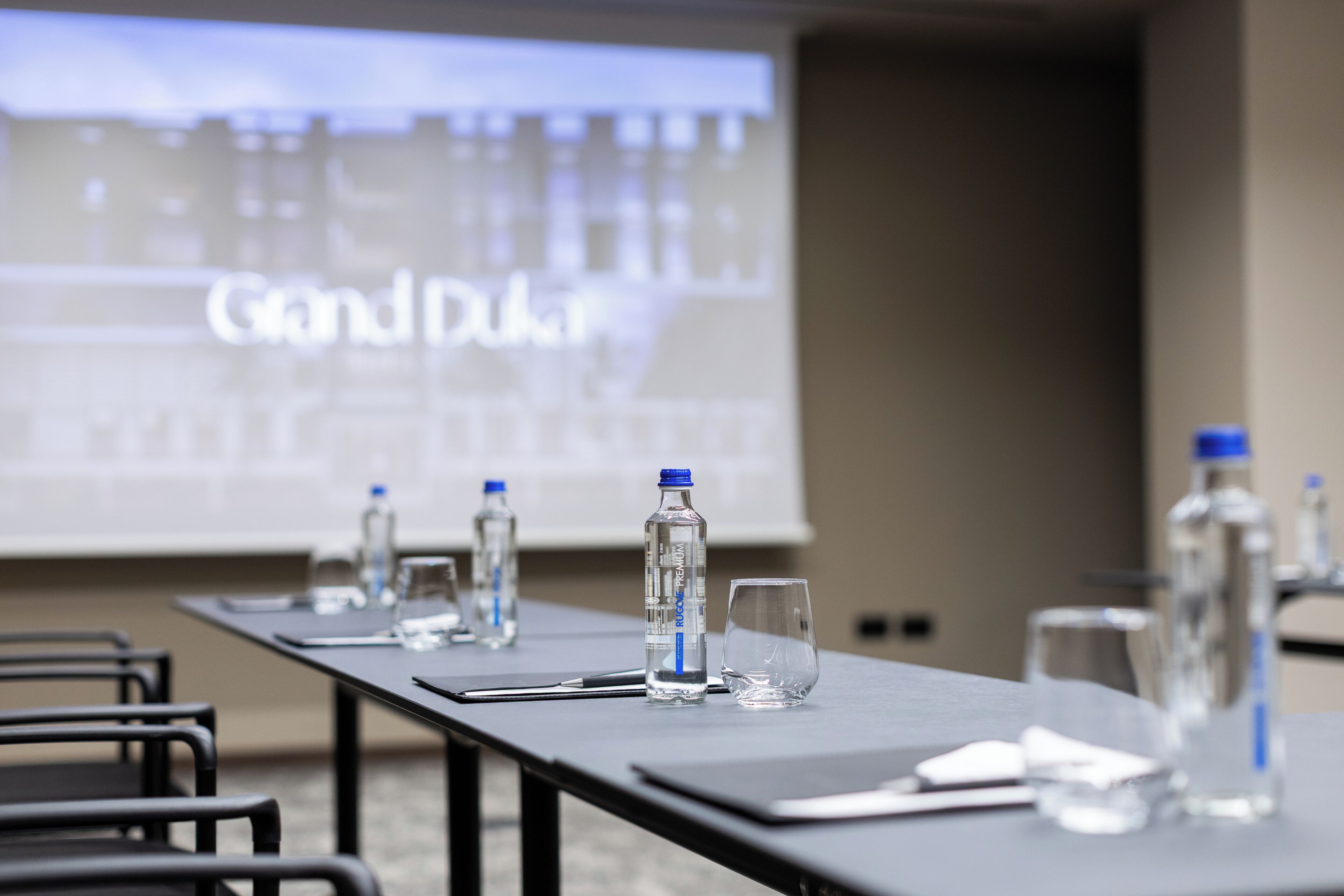 Uma sala de conferência com uma mesa comprida, garrafas de água, copos e uma tela de projetor exibindo 'Grand Duke'.
