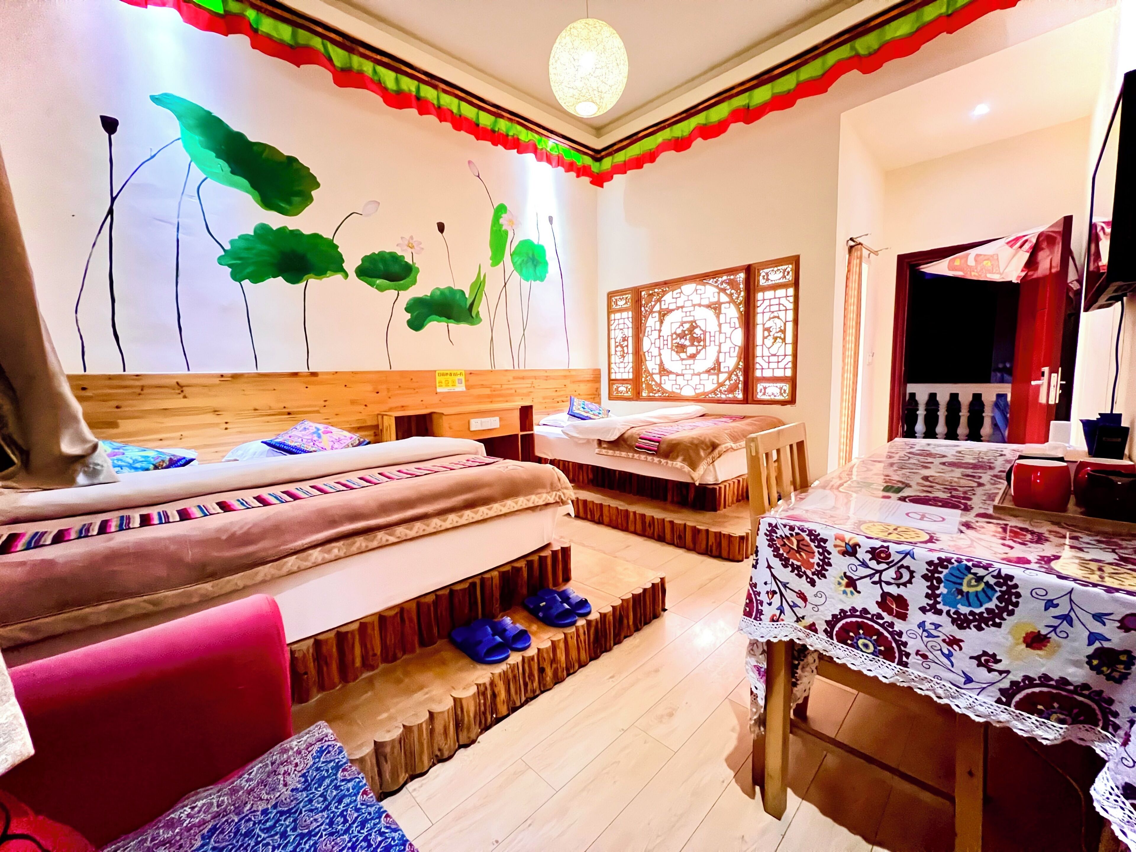 Quarto com duas camas, piso de madeira, mesa com toalha de mesa estampada e parede decorativa com motivos de lótus.