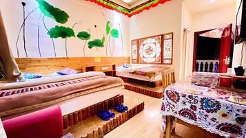 Quarto com duas camas, piso de madeira, mesa com toalha de mesa estampada e parede decorativa com motivos de lótus.