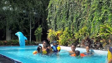 Uma piscina com pessoas na água, rodeada por vegetação exuberante e uma estátua de golfinho.