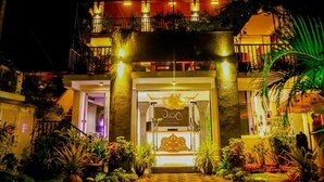 Entrada de hotel com fachada bem iluminada, varanda com grades e diversos vasos de plantas no jardim ao redor.