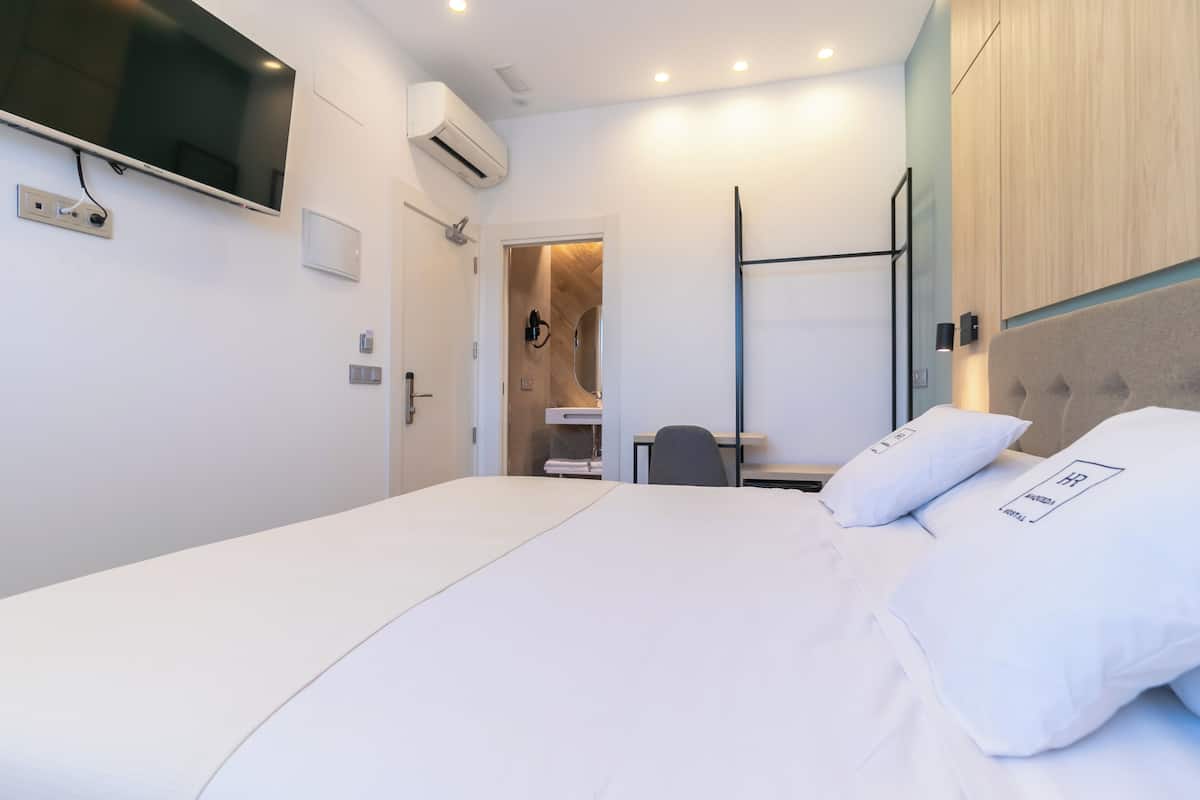 Quarto de hotel com cama, escrivaninha, cadeira, TV fixada na parede e aparelho de ar condicionado.