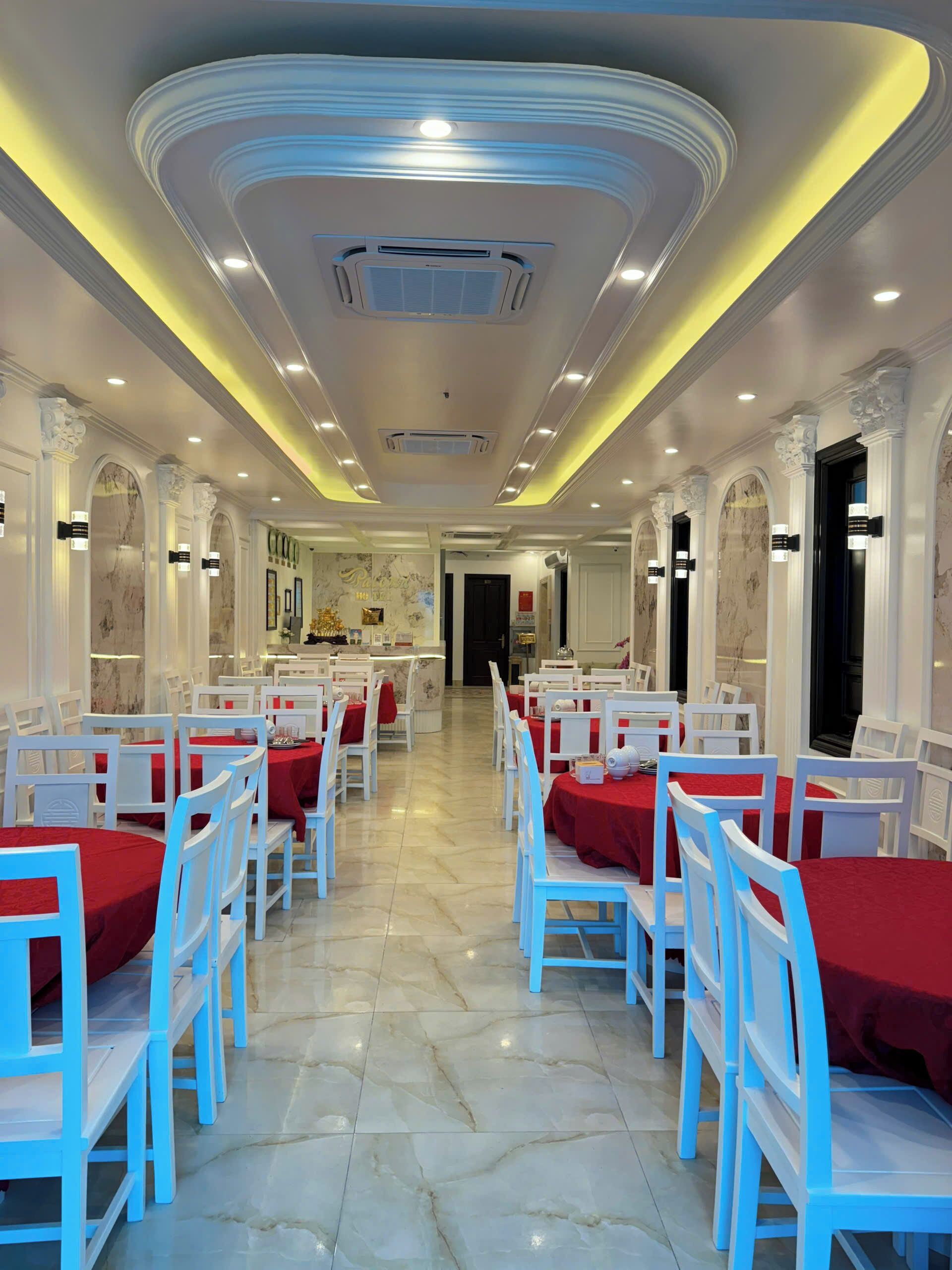 Espaço de refeições de hotel com cadeiras brancas, toalhas de mesa vermelhas, piso de mármore e design de teto moderno.