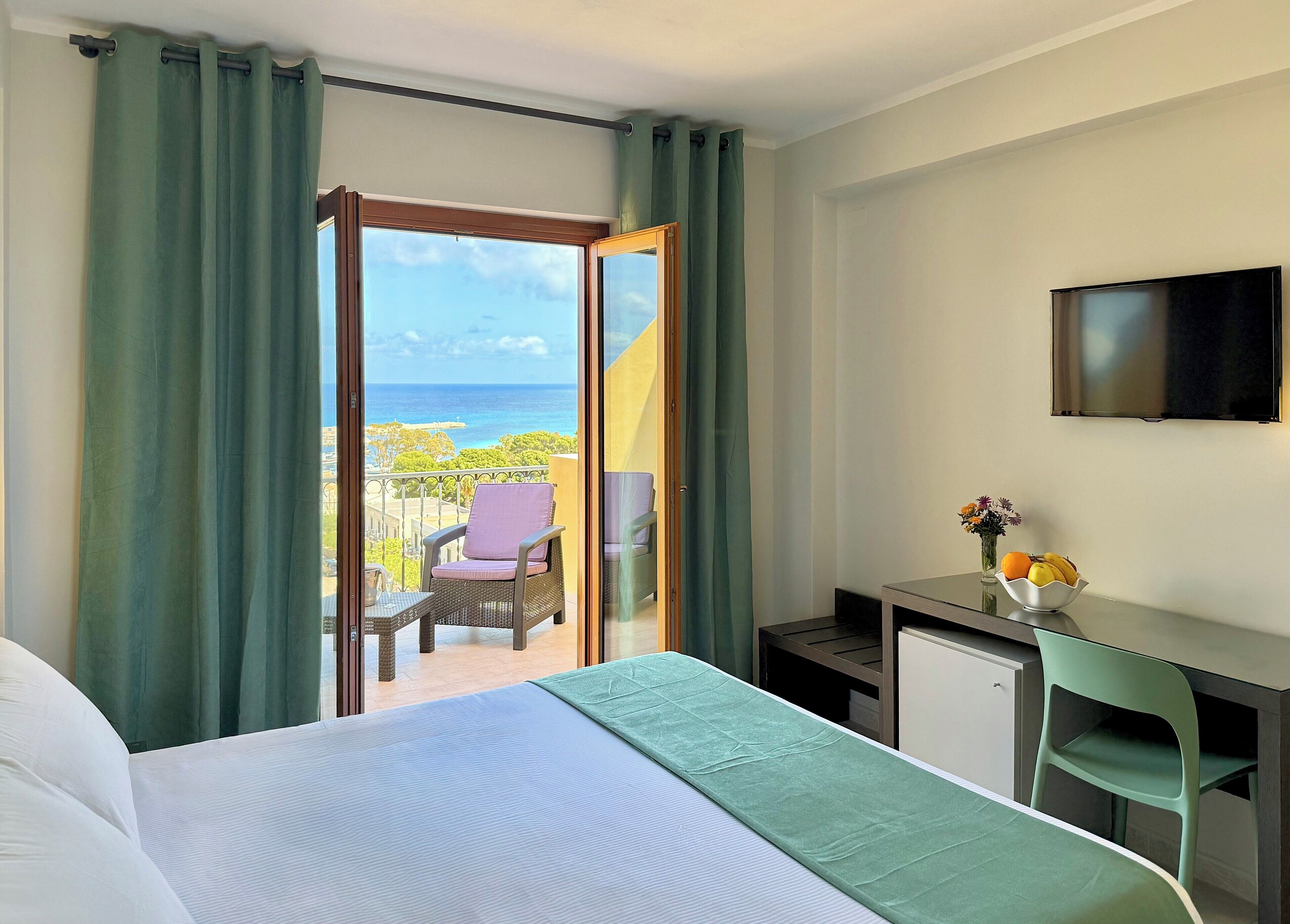 Quarto de hotel com cama, escrivaninha, cadeira, TV e varanda com vista para o mar.