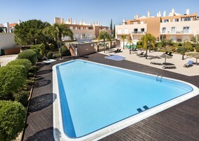 Uma piscina retangular com deck de madeira ao redor, com edifícios residenciais e palmeiras ao fundo.