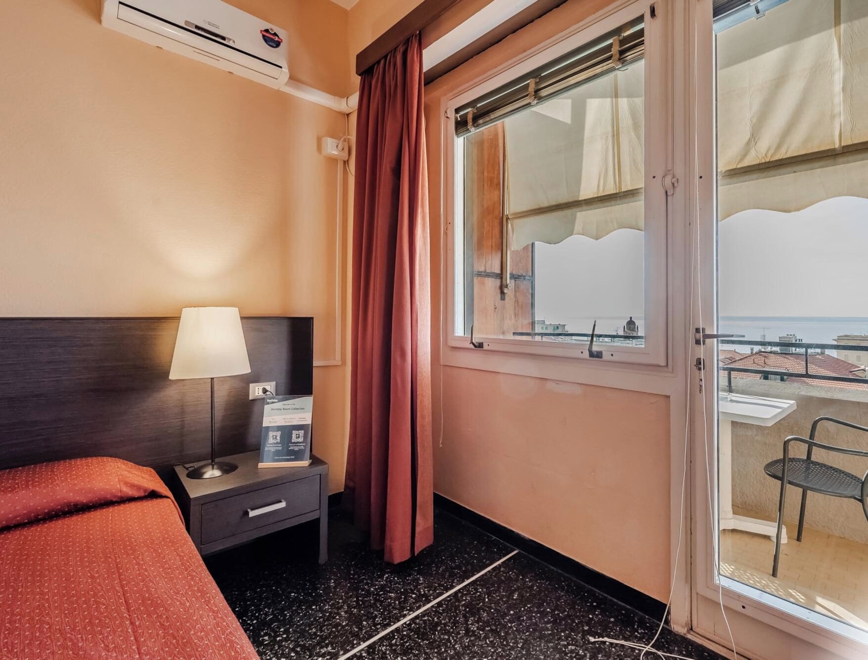 Quarto de hotel com cama, mesa de cabeceira, luminária, janela com cortinas e vista para o mar.