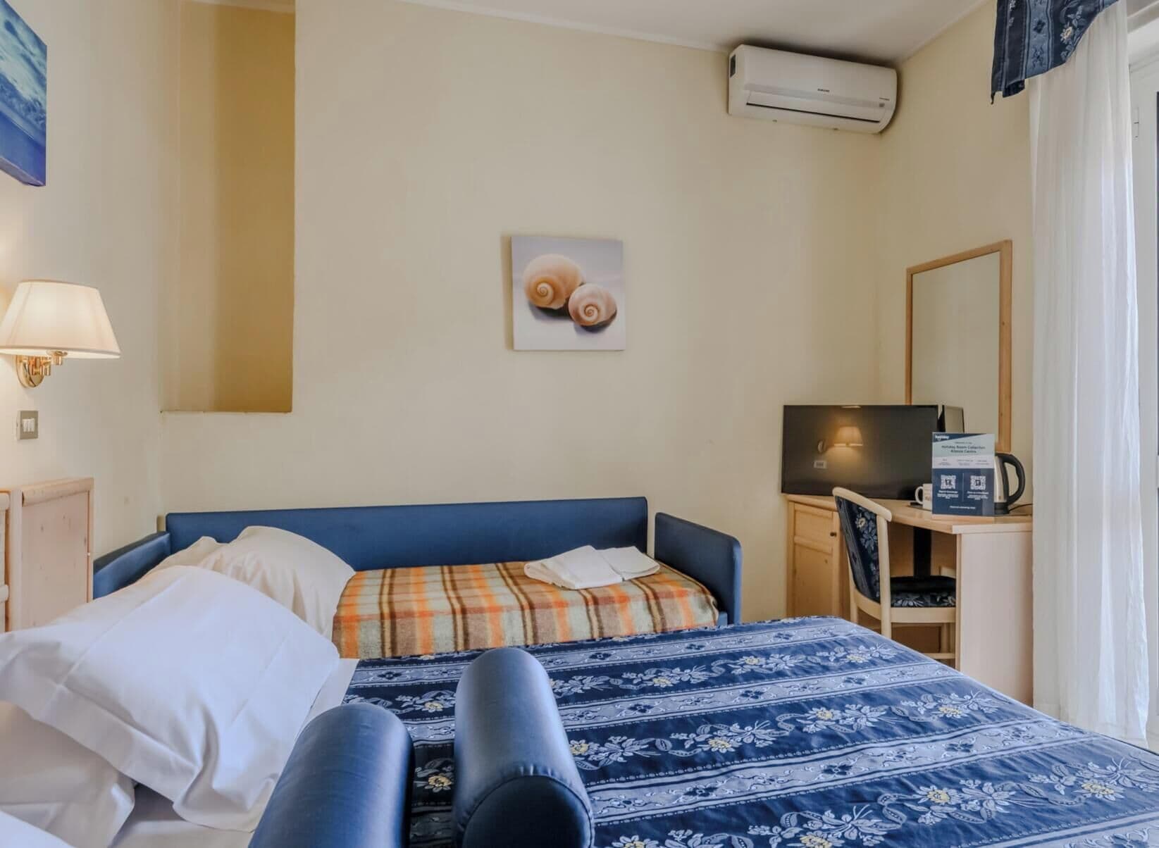Quarto de hotel com cama, escrivaninha, cadeira, ar condicionado e um quadro na parede.