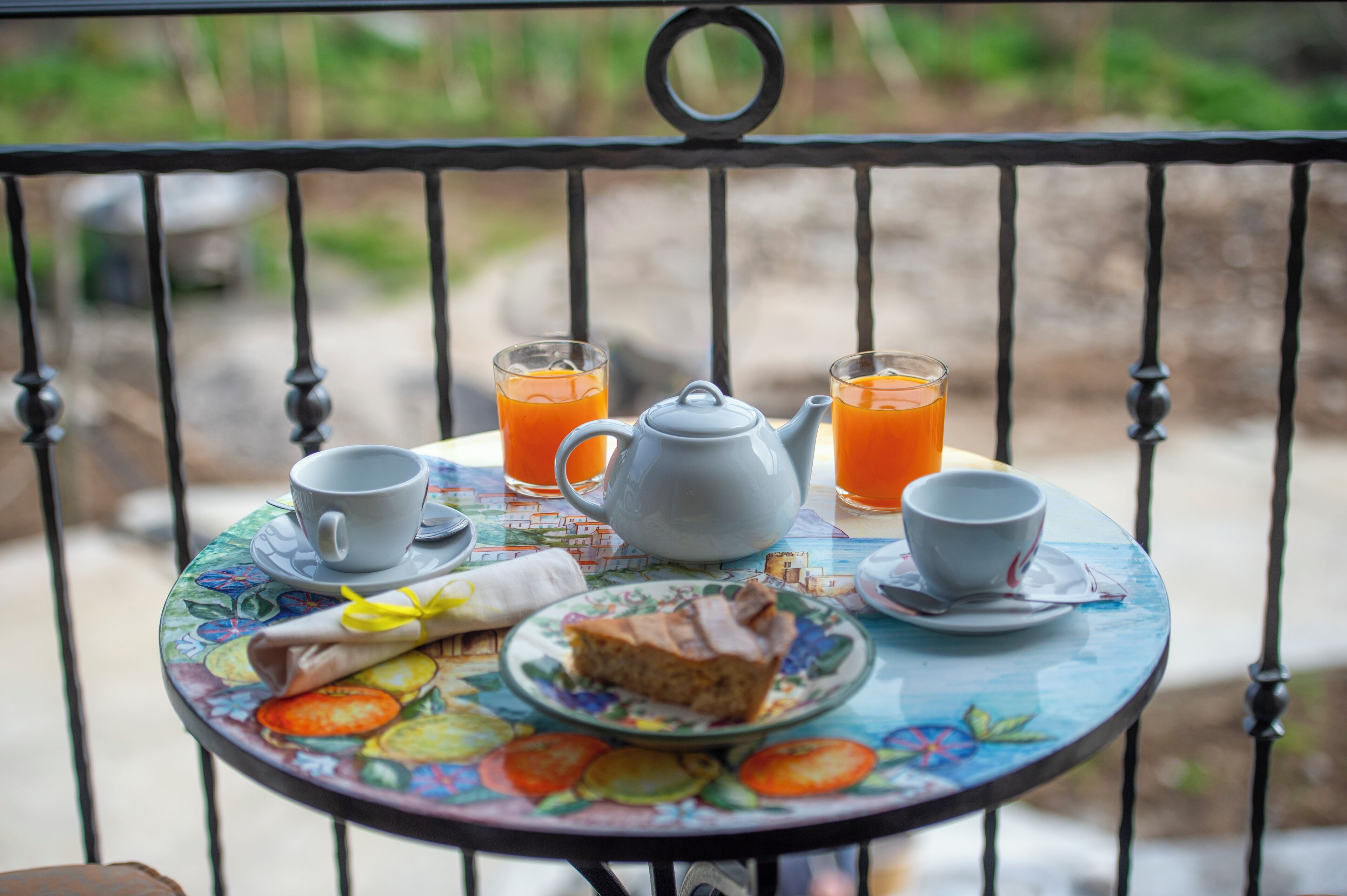 Uma mesa com um bule, duas xícaras, um copo de sumo de laranja e uma fatia de bolo sobre um tecido de mesa estampado com flores.