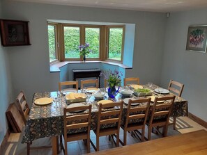 Uma sala de jantar com janela panorâmica, cadeiras de madeira, toalha de mesa florida e um vaso com flores.