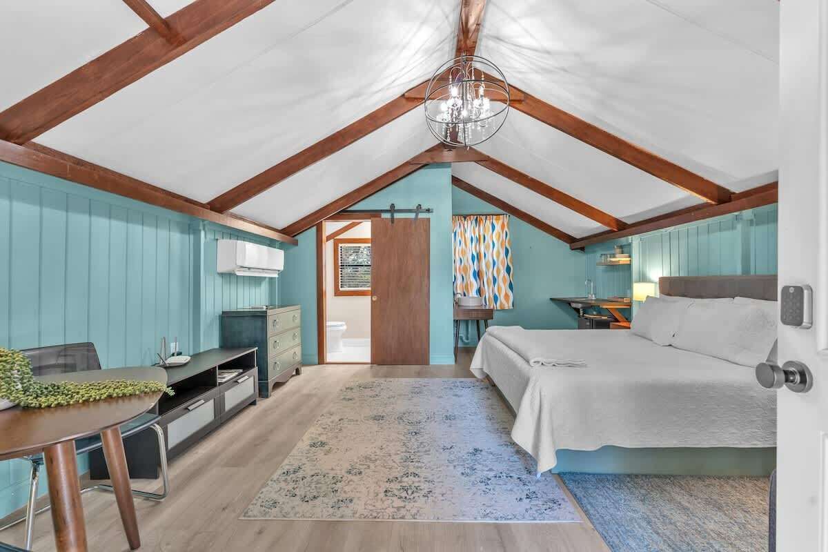 Um quarto com uma cama, uma escrivaninha e uma cadeira. Há uma porta que dá para outro cômodo.