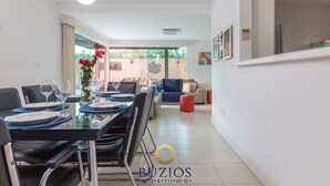 Uma área de refeições com mesa de vidro, cadeiras pretas e um banquinho vermelho. A mesa está posta com pratos, copos e um vaso de flores.