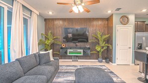 Uma sala de estar moderna com um sofá cinza, um banquinho cinza, uma TV de tela plana e um ventilador de teto.