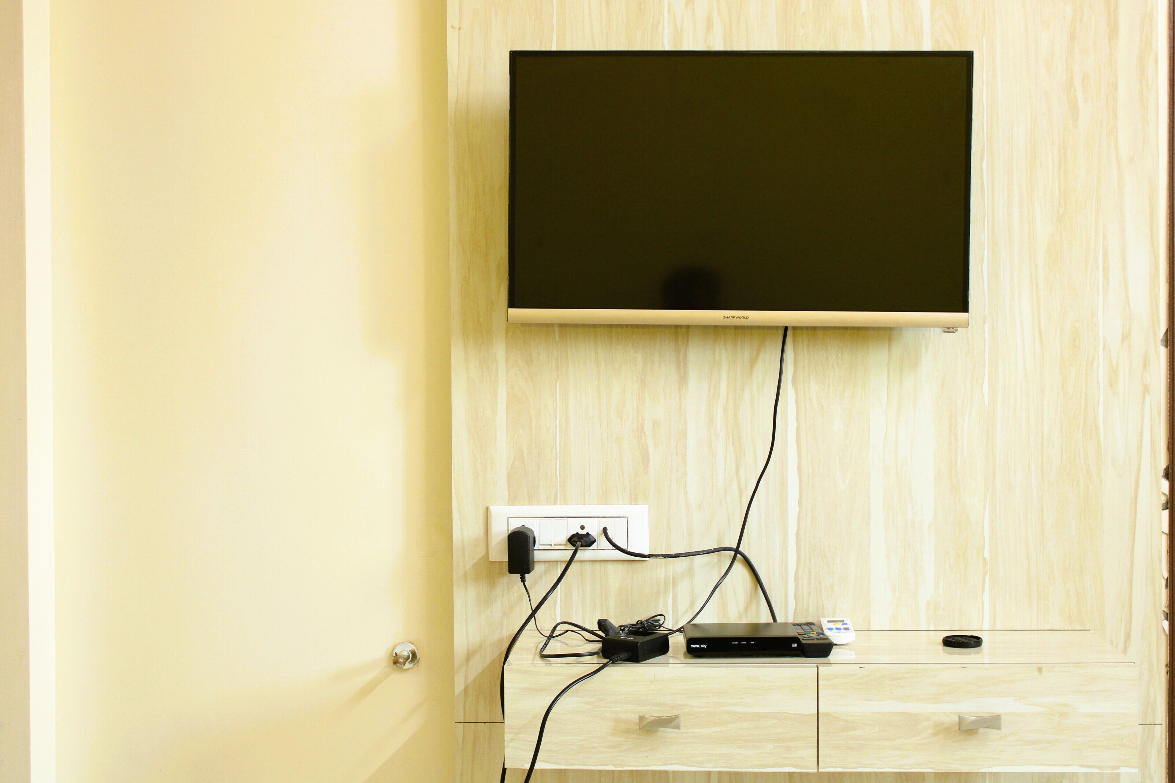 Uma televisão fixada na parede, com uma régua de tomadas e um controle remoto.