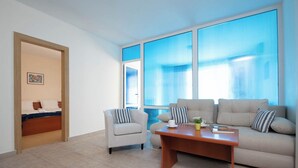 Uma sala de estar moderna com um sofá, poltrona e mesa de centro. Há uma cama no quarto vizinho, e janelas amplas proporcionam iluminação natural.