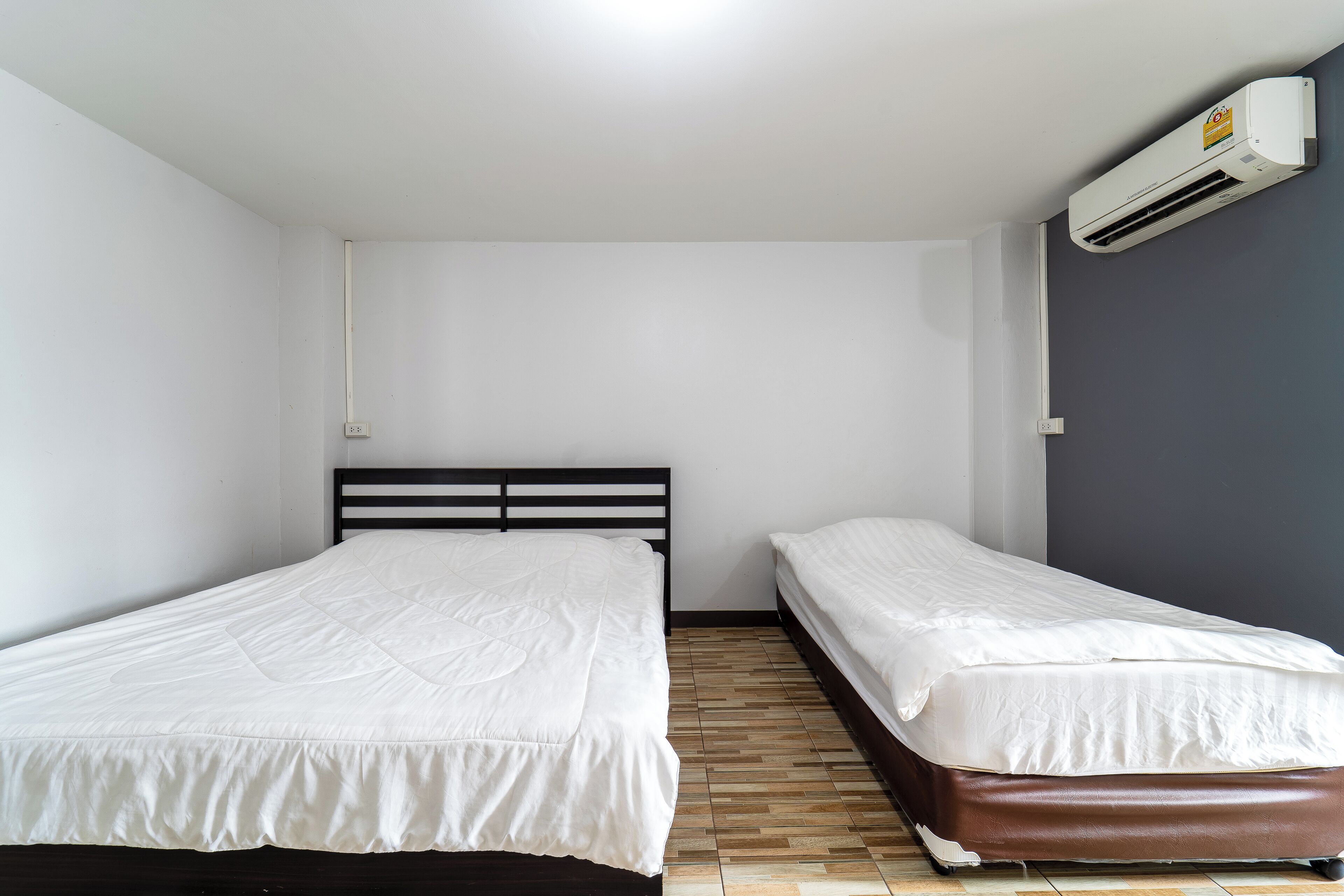 Quarto pequeno e limpo com duas camas de solteiro, piso de madeira e um ar condicionado na parede.